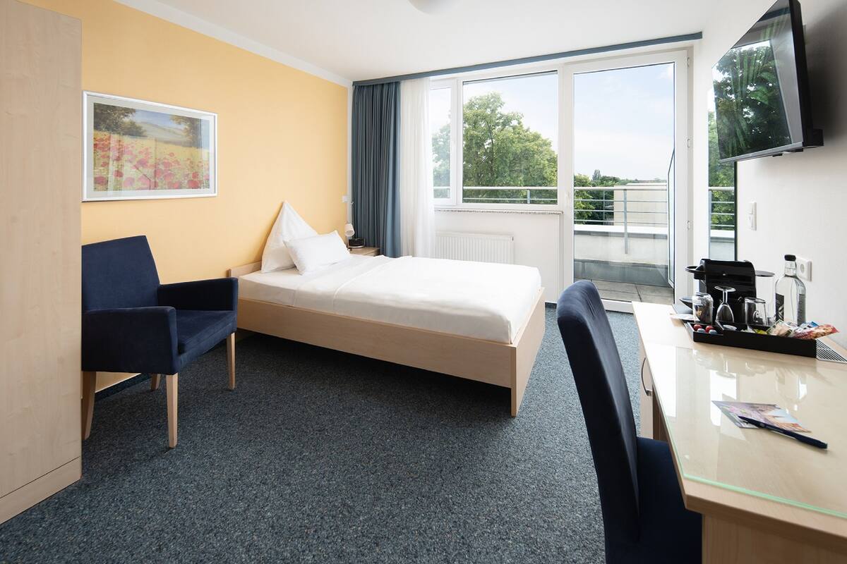 Quarto de hotel com uma cama, escrivaninha, cadeira, televisão e janela com vista para o verde.