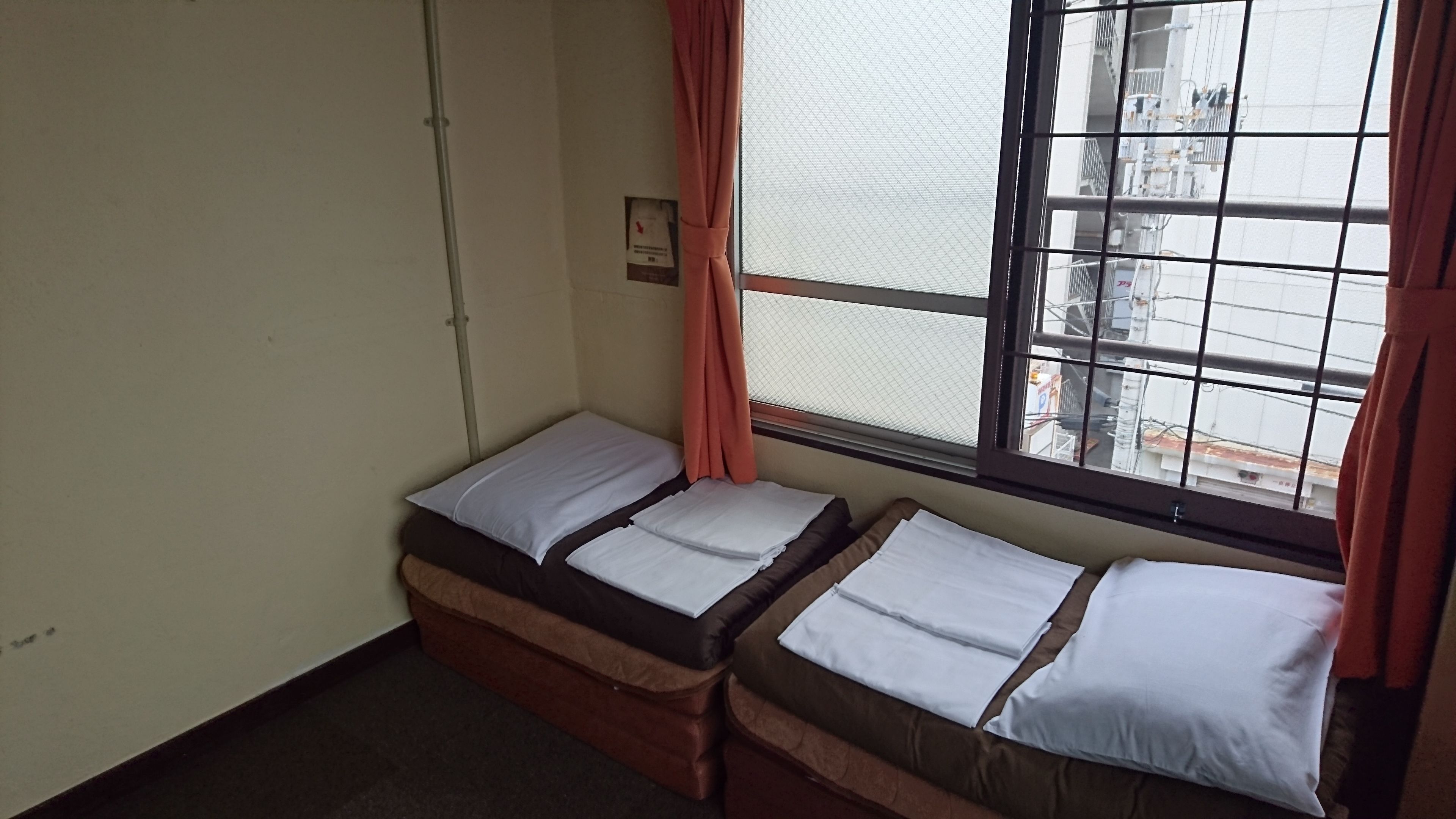 Quarto com duas camas de solteiro, uma janela com vista para um edifício e uma cortina.