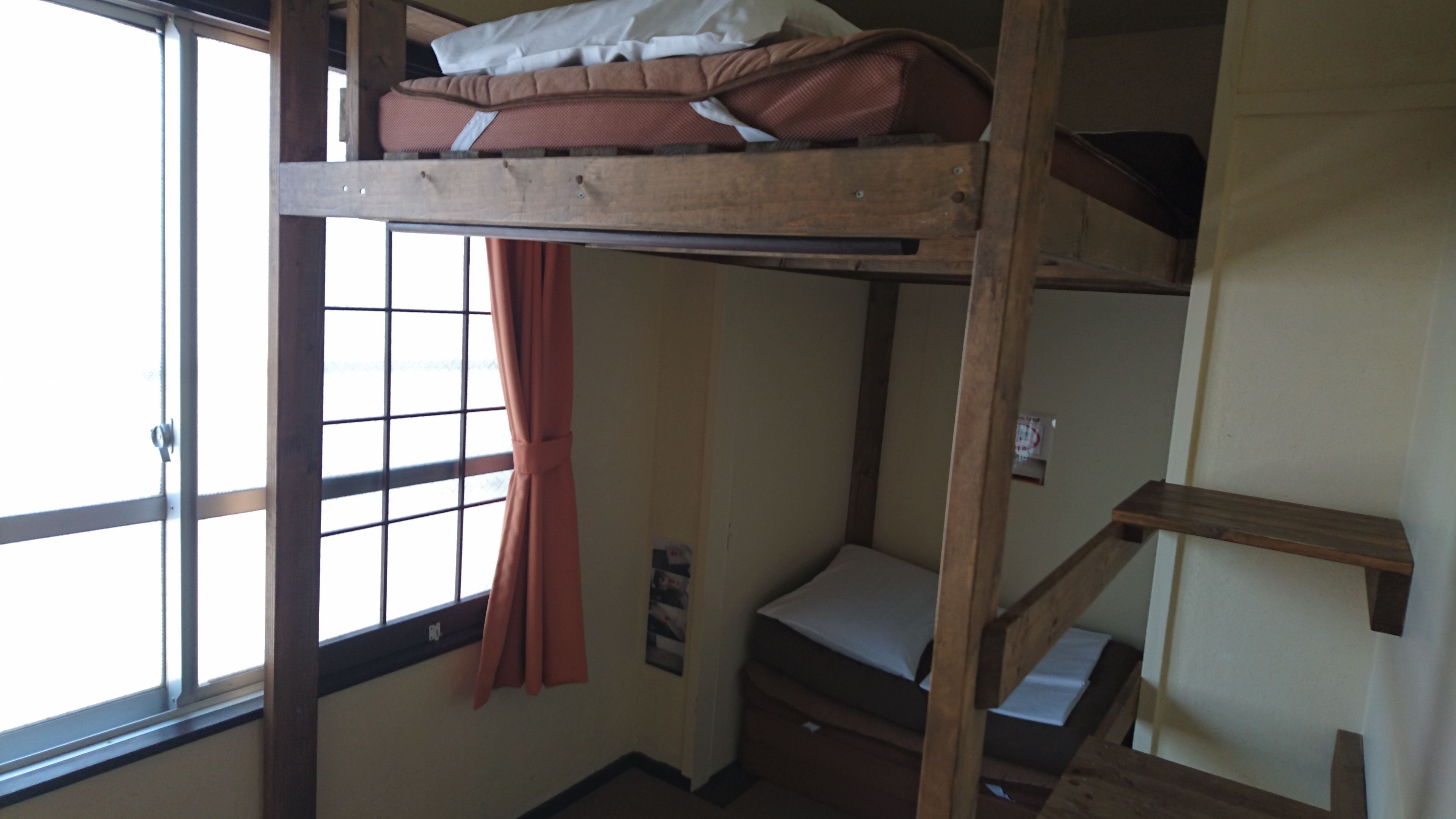Uma cama de solteiro com colchão e almofadas no topo, uma pequena mesa de cabeceira e uma janela com cortinas.