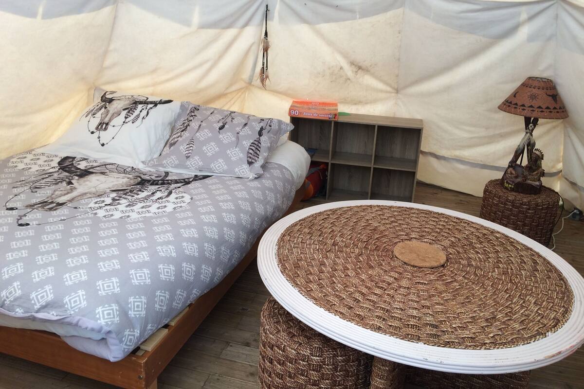 Um quarto com tenda, uma cama, uma mesa redonda de palha e um abajur.