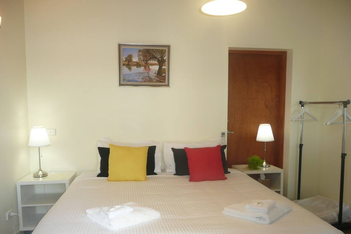 Quarto de hotel com cama, almofadas, mesa de cabeceira, luminária, quadro na parede e uma porta.
