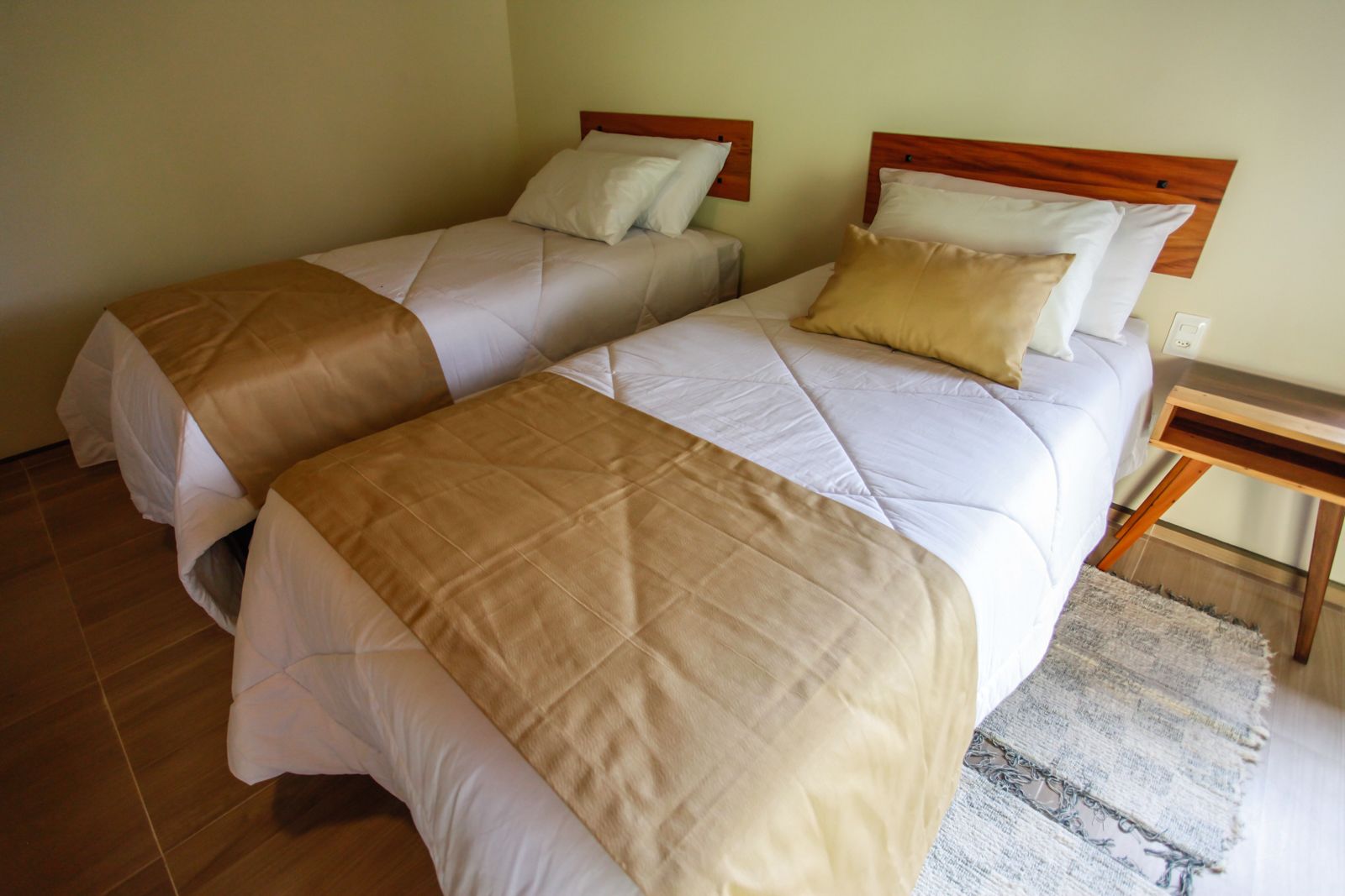 Quarto com duas camas, cada uma com cabeceira de madeira, roupa de cama branca e manta bege. Há uma pequena mesa de cabeceira de madeira com uma luminária ao lado direito.