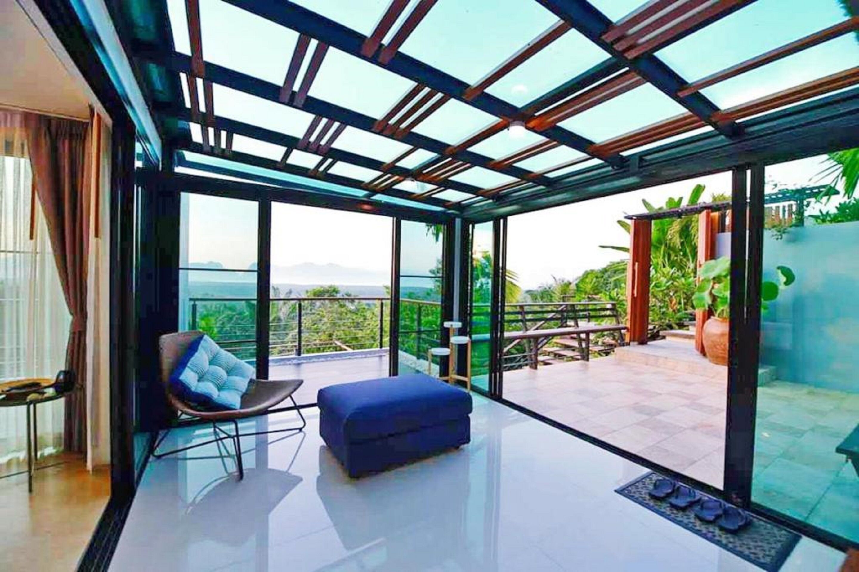 Sala de estar moderna com teto de vidro, sofá azul e vista para área verde e montanhas.