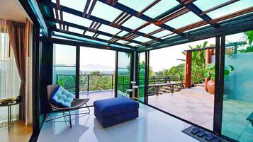 Sala de estar moderna com teto de vidro, sofá azul e vista para área verde e montanhas.