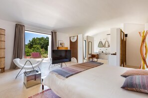 Quarto moderno com uma cama grande, televisão de tela plana, uma cadeira branca e vista para o verde lá fora.