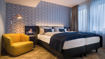 Quarto de hotel com cama, escrivaninha, cadeira, abajur e papel de parede estampado.