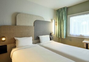 Quarto de hotel com duas camas, cabeceira com padrão texturizado, mesa de cabeceira com abajur e janela com cortinas.