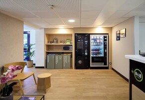 Uma área de recepção de hotel com piso de madeira, balcão de recepção, máquina de vendas automática e um espaço de espera com uma cadeira e uma mesa pequena.