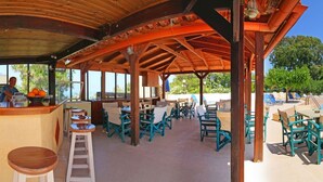 Área de piscina ao ar livre com espreguiçadeiras e mesas, um bar com banquetas e uma área de estar coberta.