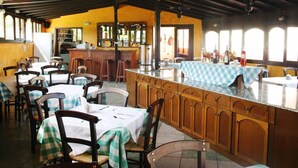 Restaurante com mesas e cadeiras de madeira, área de bar e cozinha visível ao fundo.