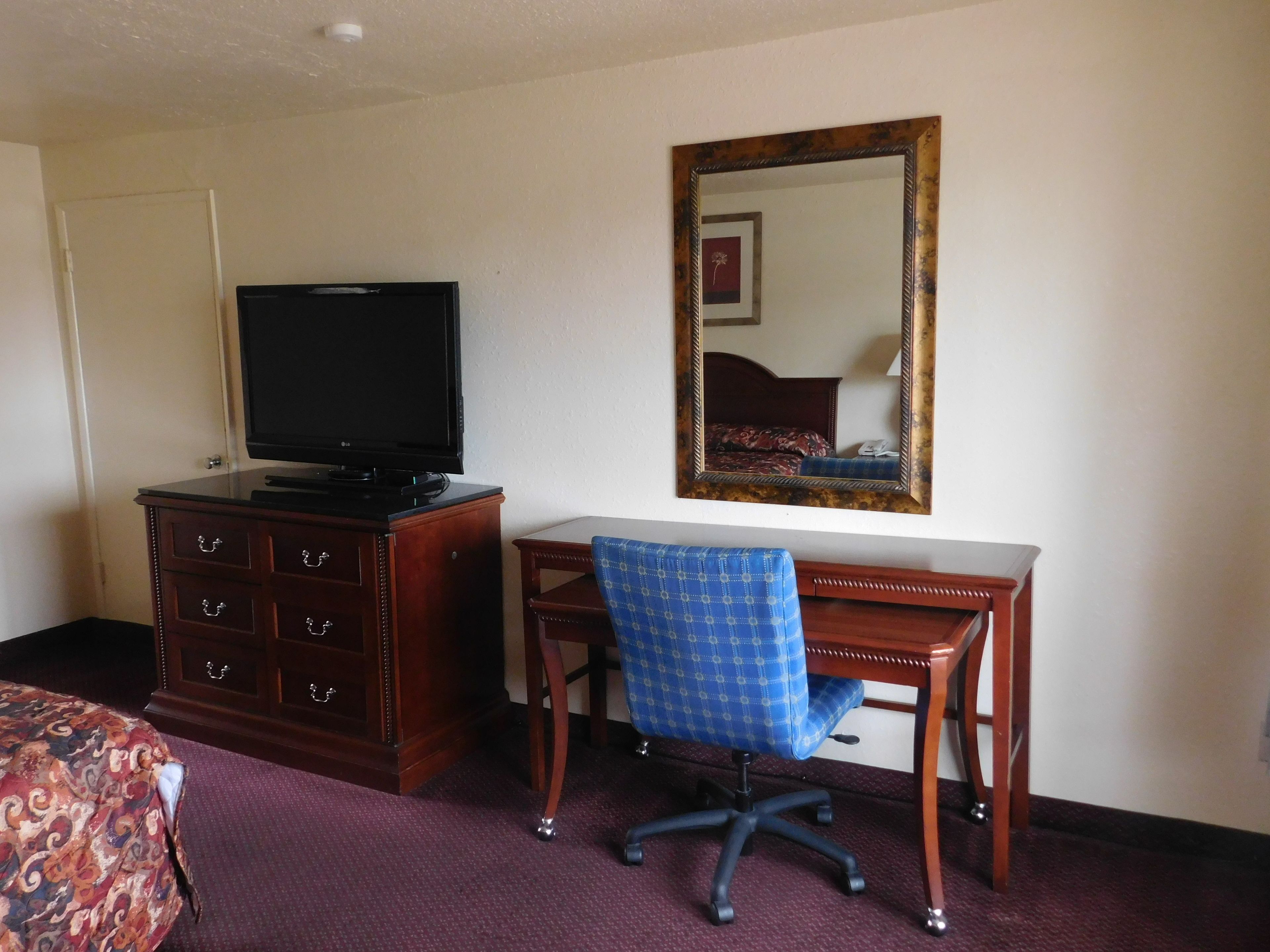 Quarto de hotel com cômoda de madeira, televisão, escrivaninha com cadeira e um espelho grande na parede.