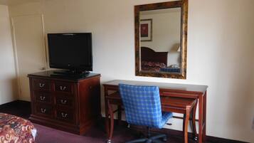 Quarto de hotel com cômoda de madeira, televisão, escrivaninha com cadeira e um espelho grande na parede.