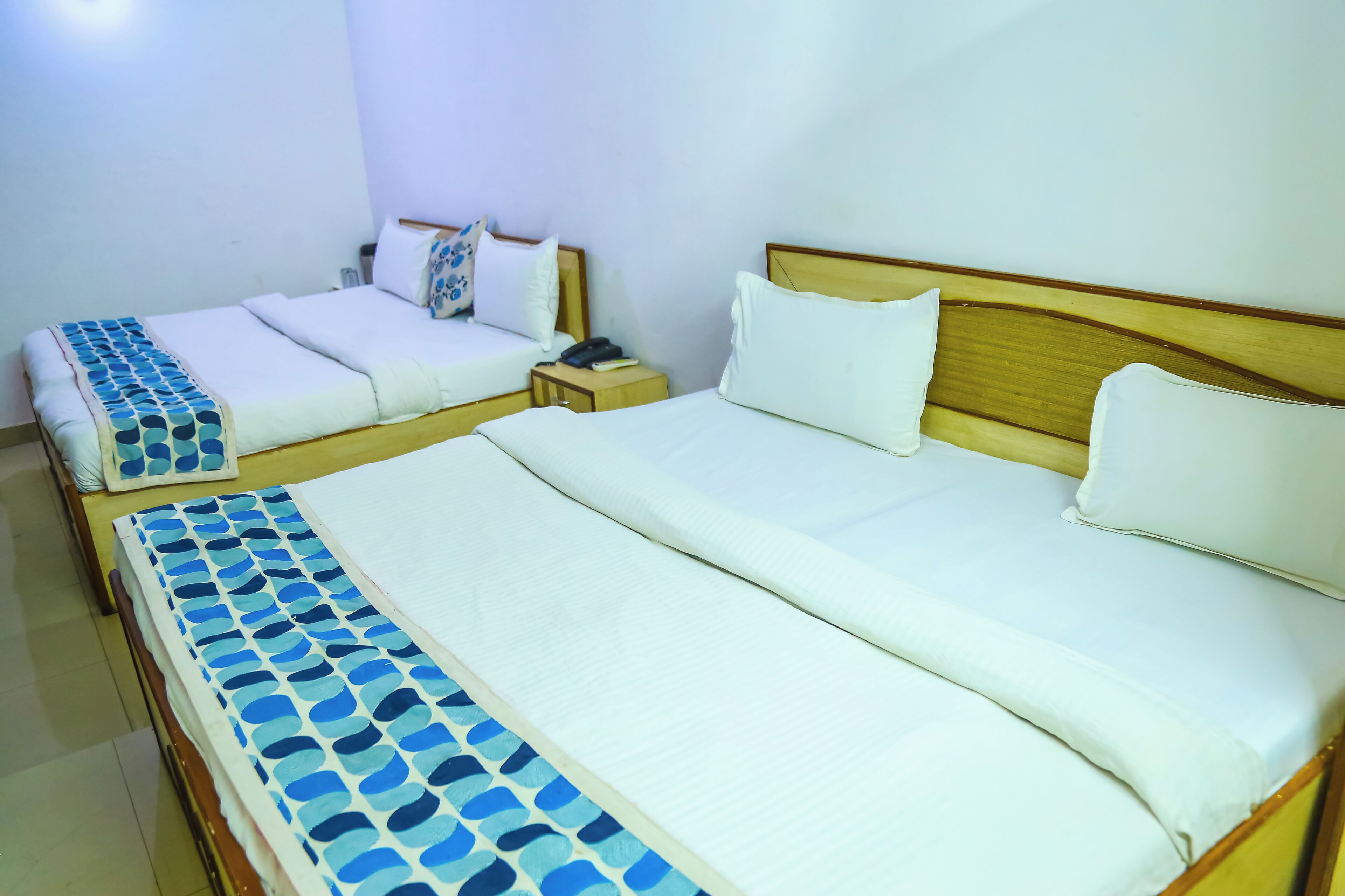 Quarto de hotel com duas camas, cada uma com roupa de cama branca e cabeceira de madeira. Há uma pequena mesa de cabeceira com uma luminária e um telefone.