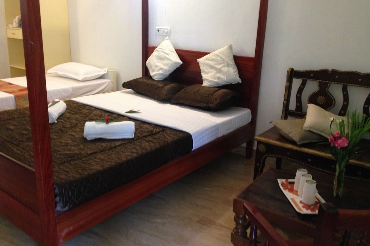 Uma cama de dossel com colcha marrom, lençóis brancos e duas almofadas. Uma mesinha com bandeja de artigos de higiene pessoal e um vaso com flores fica ao lado da cama.