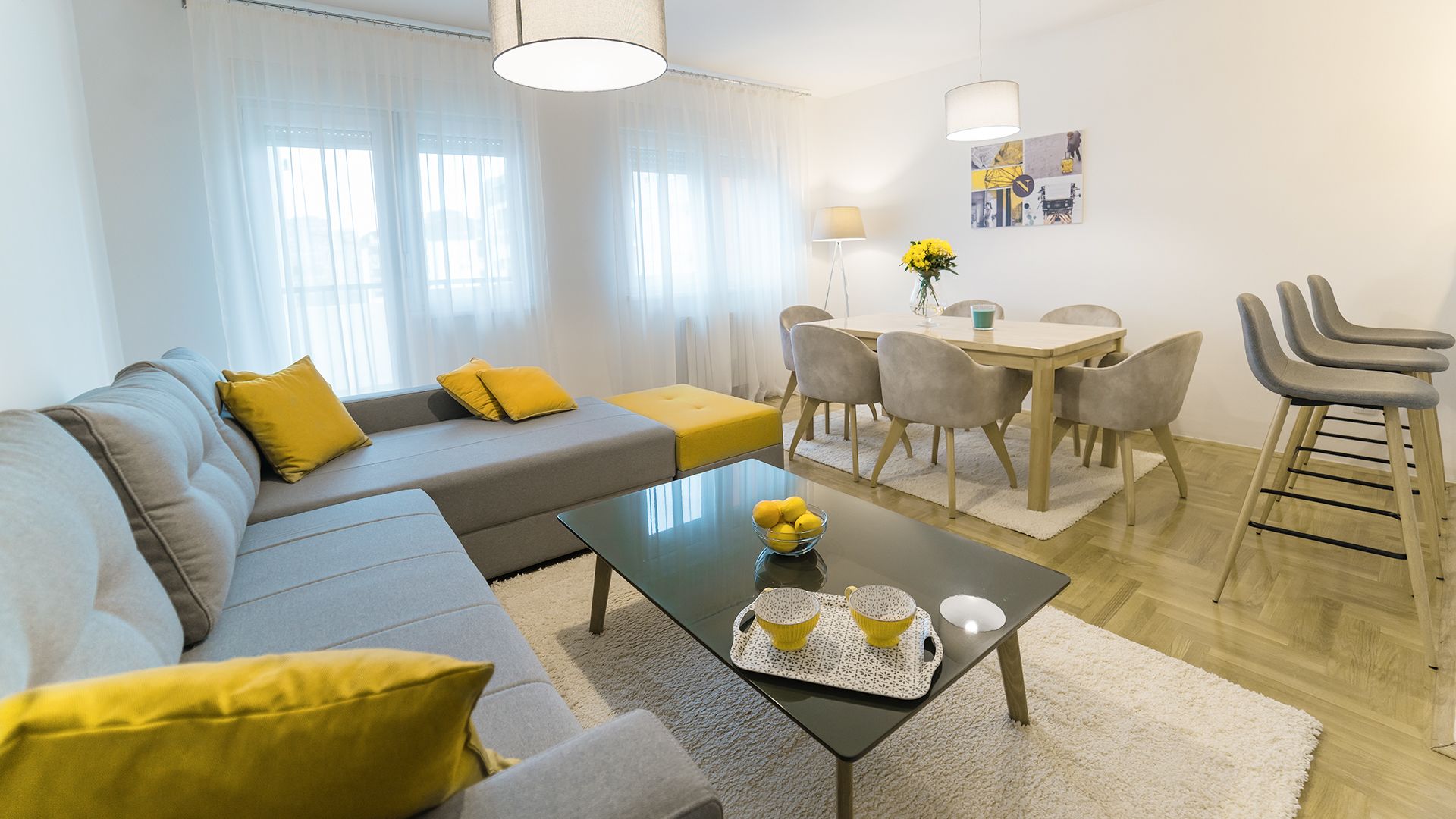 Uma sala de estar moderna com um sofá cinzento, almofadas amarelas, uma mesa de centro de vidro e uma área de jantar com uma mesa e cadeiras brancas.