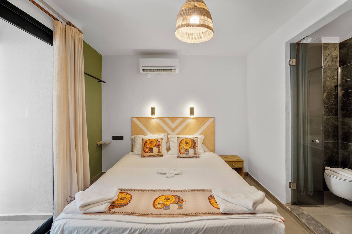 Quarto de hotel com cama, ar condicionado, chuveiro e luminária pendente.