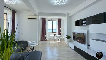 Uma sala de estar moderna com sofá, mesa de jantar com cadeiras, TV na parede e vista para o mar pela janela.