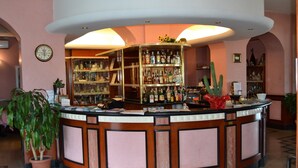 Uma área de bar com balcão circular, prateleiras com garrafas e um relógio na parede.