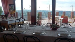 Uma sala de jantar com mesas postas para refeição, grandes janelas com vista para a paisagem urbana e plantas em vasos sobre as mesas.