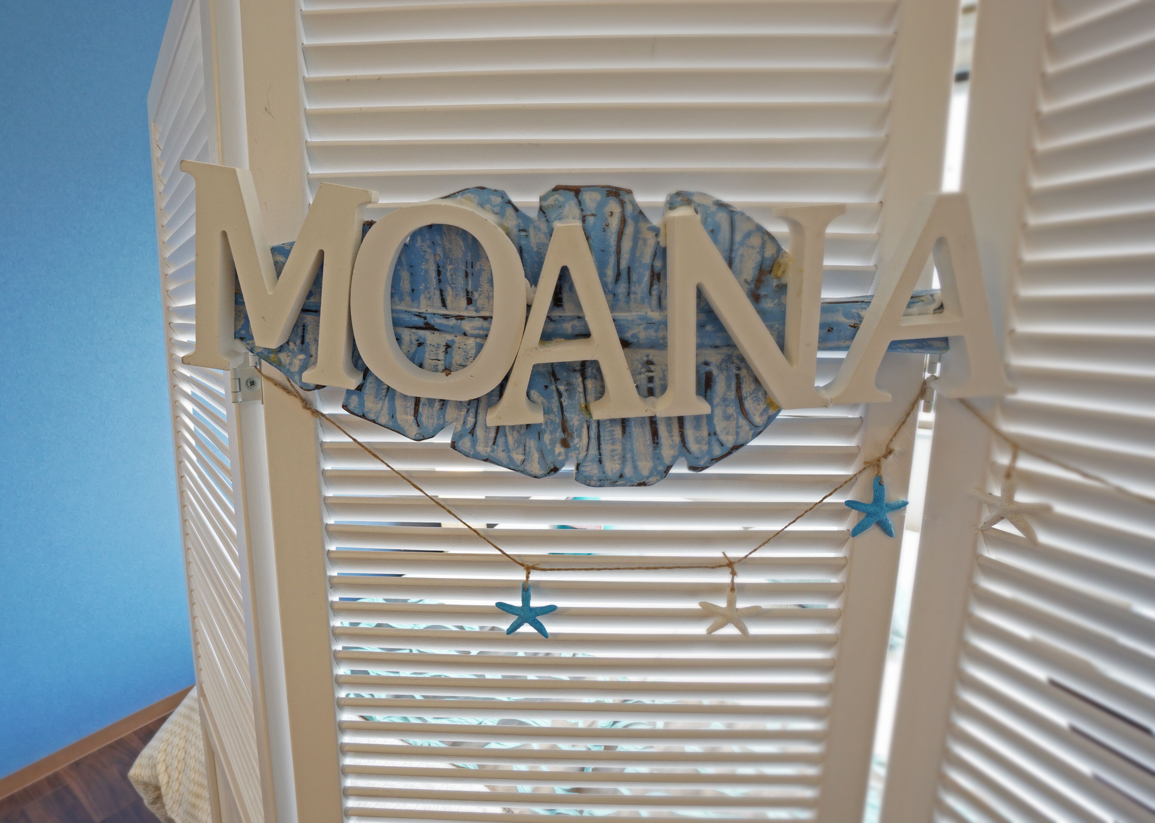 Uma placa decorativa com o nome 'MOANA' em letras grandes, pendurada em frente a uma janela com persianas horizontais.