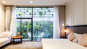 Quarto de hotel com uma grande porta de vidro deslizante para um jardim, uma cama com almofadas estampadas, um sofá branco e uma pequena mesa com um vaso.