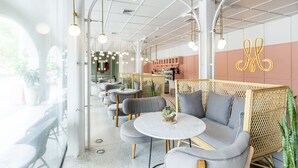 Um café moderno com mesas redondas e assentos acolchoados cinzentos, com teto branco e um grande letreiro dourado 'HH' na parede.