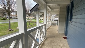 Um espaço ao ar livre coberto, com piso de azulejos, grades brancas e vista para uma estrada e edifícios ao fundo.