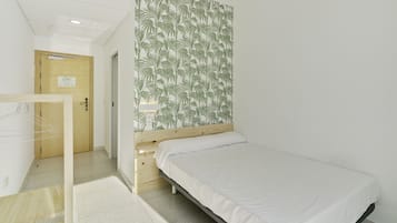 Um quarto pequeno e bem organizado, com uma cama de solteiro, cabeceira de madeira e parede com padrão de folhagem tropical.