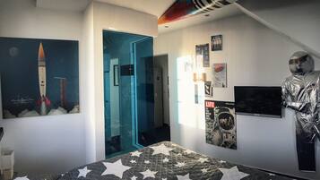 Um quarto com uma cama coberta por um edredom com padrão de estrelas, um cartaz de lançamento de foguete e diversas decorações com tema espacial nas paredes.