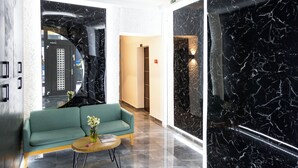 Um lobby com sofá verde, uma mesa de madeira redonda com um vaso e paredes de mármore preto.