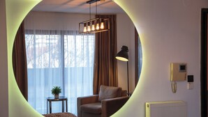 Sala de estar moderna com um espelho redondo refletindo o interior. Possui uma poltrona confortável, uma mesinha de apoio com uma planta, um abajur e um radiador.