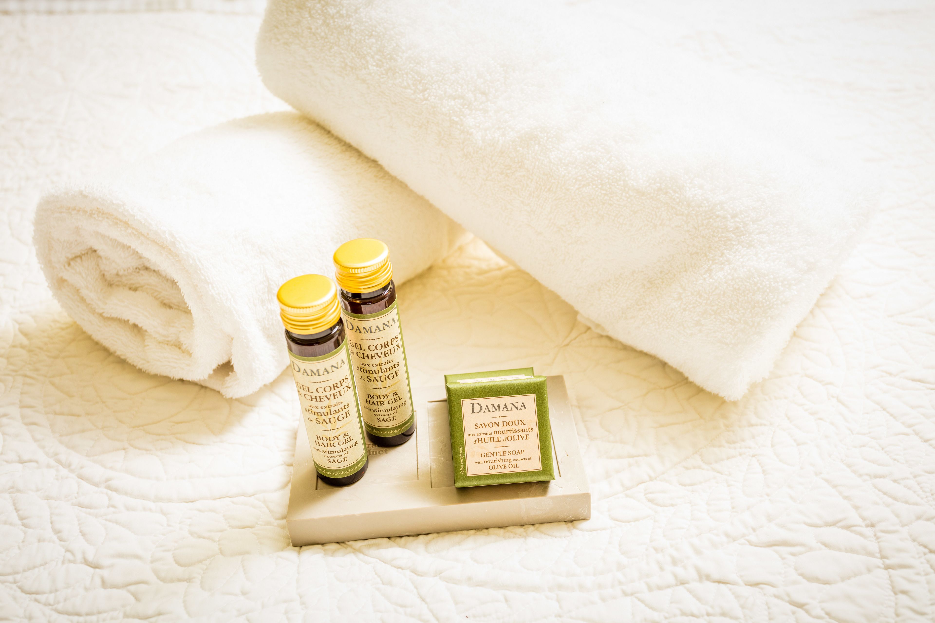 Uma toalha branca dobrada, dois frascos de produtos Damana para corpo e cabelo, e uma pequena caixa verde com sabonete Damana sobre uma superfície branca texturizada.
