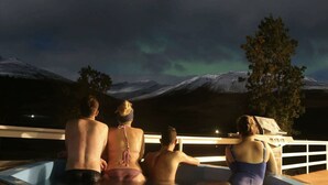 Um grupo de pessoas sentadas perto de uma piscina à noite, com vista para montanhas cobertas de neve e auroras boreais.