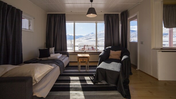 Quarto com duas camas, uma mesa pequena e uma cadeira. Janelas amplas com vista para montanhas.