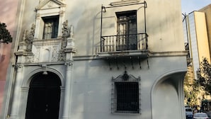 Um edifício histórico com entrada central, varanda com grades elaboradas e um relógio acima da entrada.