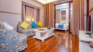 Quarto de hotel com sofá floral, uma mesa de centro branca e cama com manta azul.