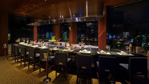 Um restaurante com uma longa mesa de jantar, várias cadeiras e vista para a cidade através de grandes janelas.