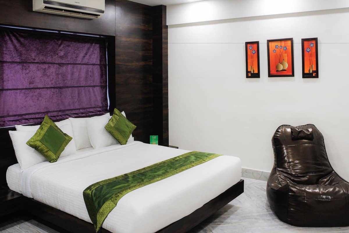 Quarto de hotel com uma cama, cabeceira roxa, almofadas verdes, uma poltrona de couro marrom e três obras de arte emolduradas na parede.
