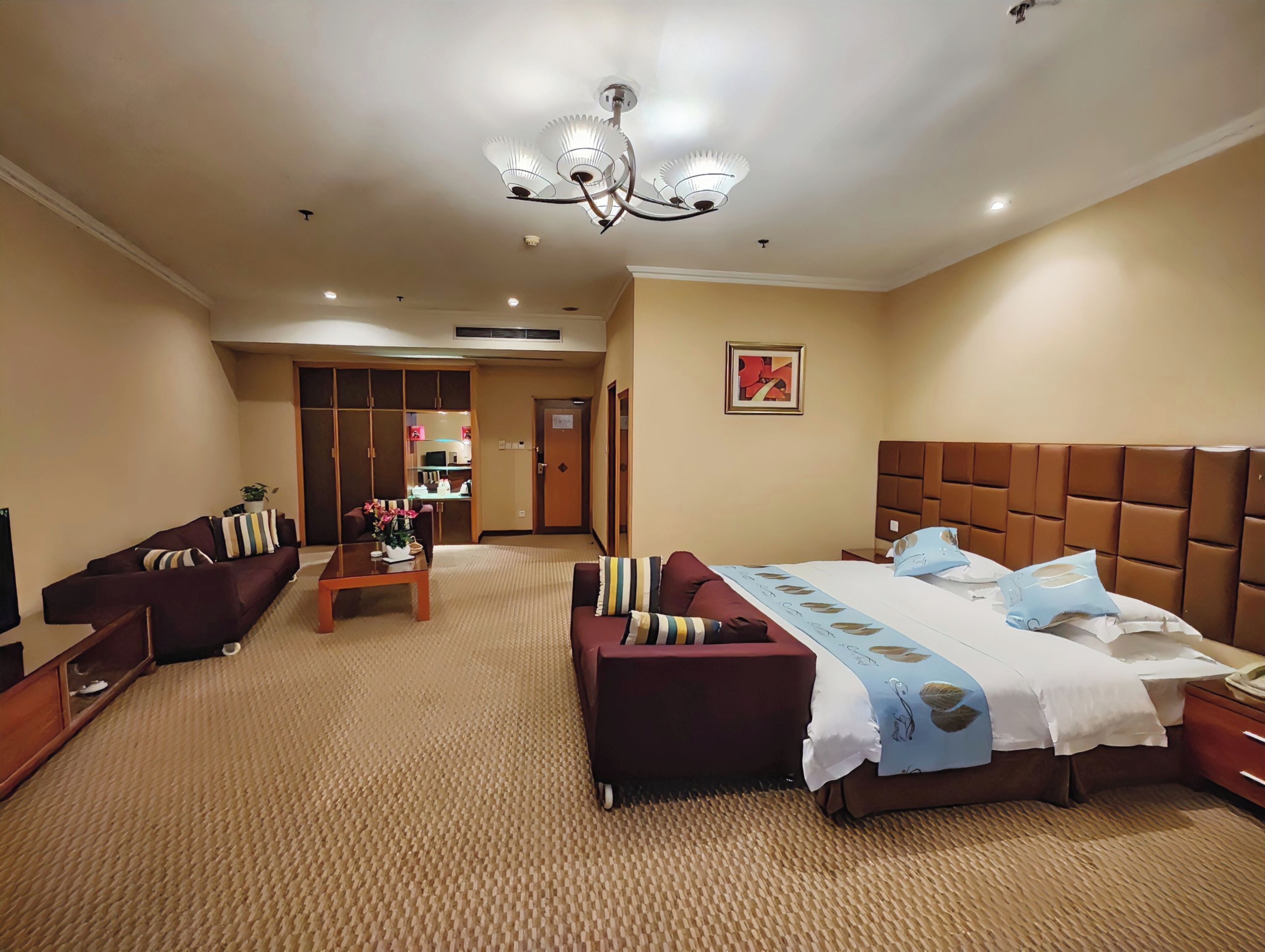 Quarto de hotel com uma cama grande, um sofá, uma pequena mesa com flores, um guarda-roupa e um candeeiro no teto.