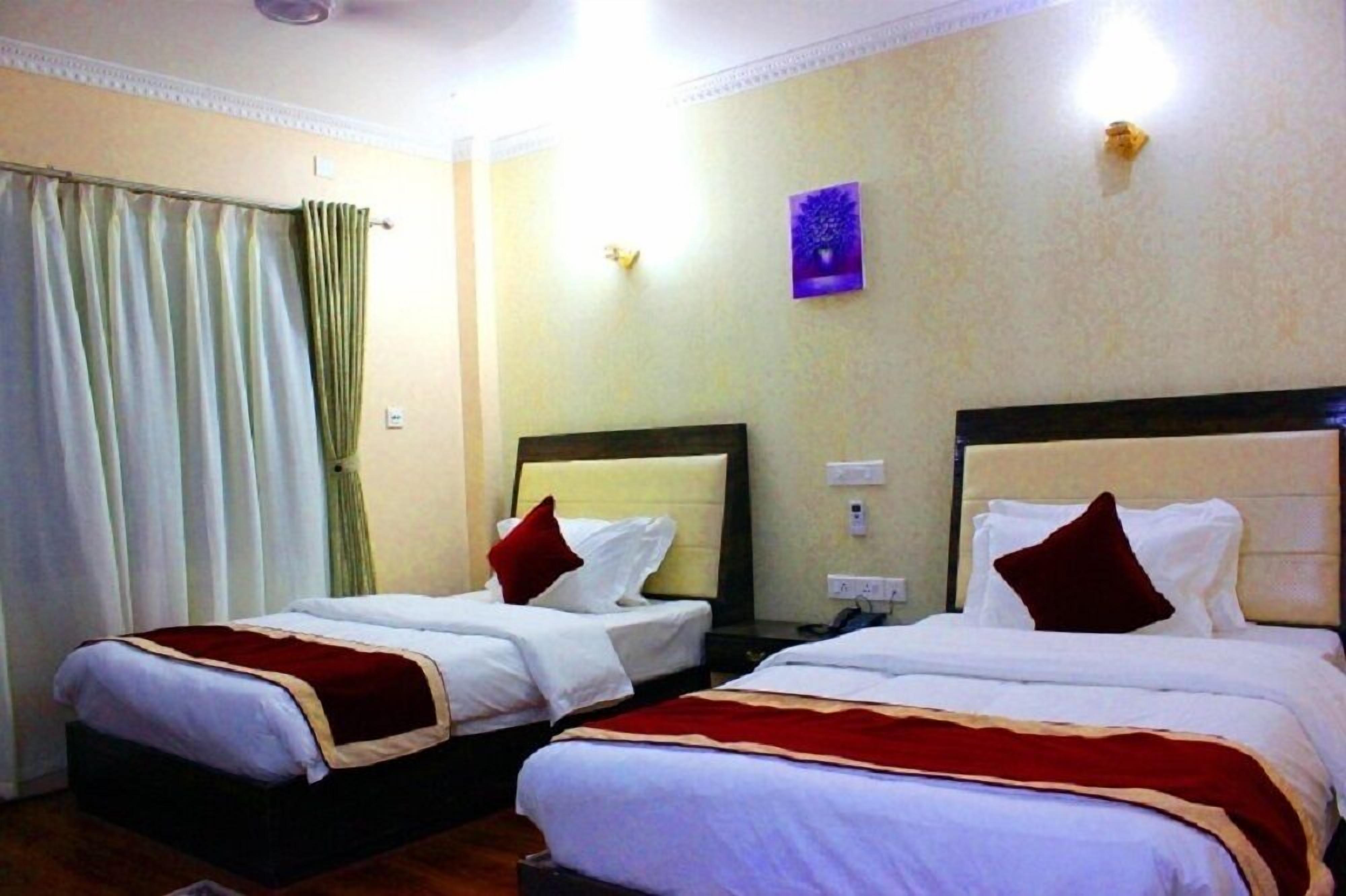 Quarto de hotel com duas camas de solteiro, cada uma com uma manta vermelha e roupa de cama branca. Há duas mesas de cabeceira, uma luminária fixada na parede e uma janela com cortinas.
