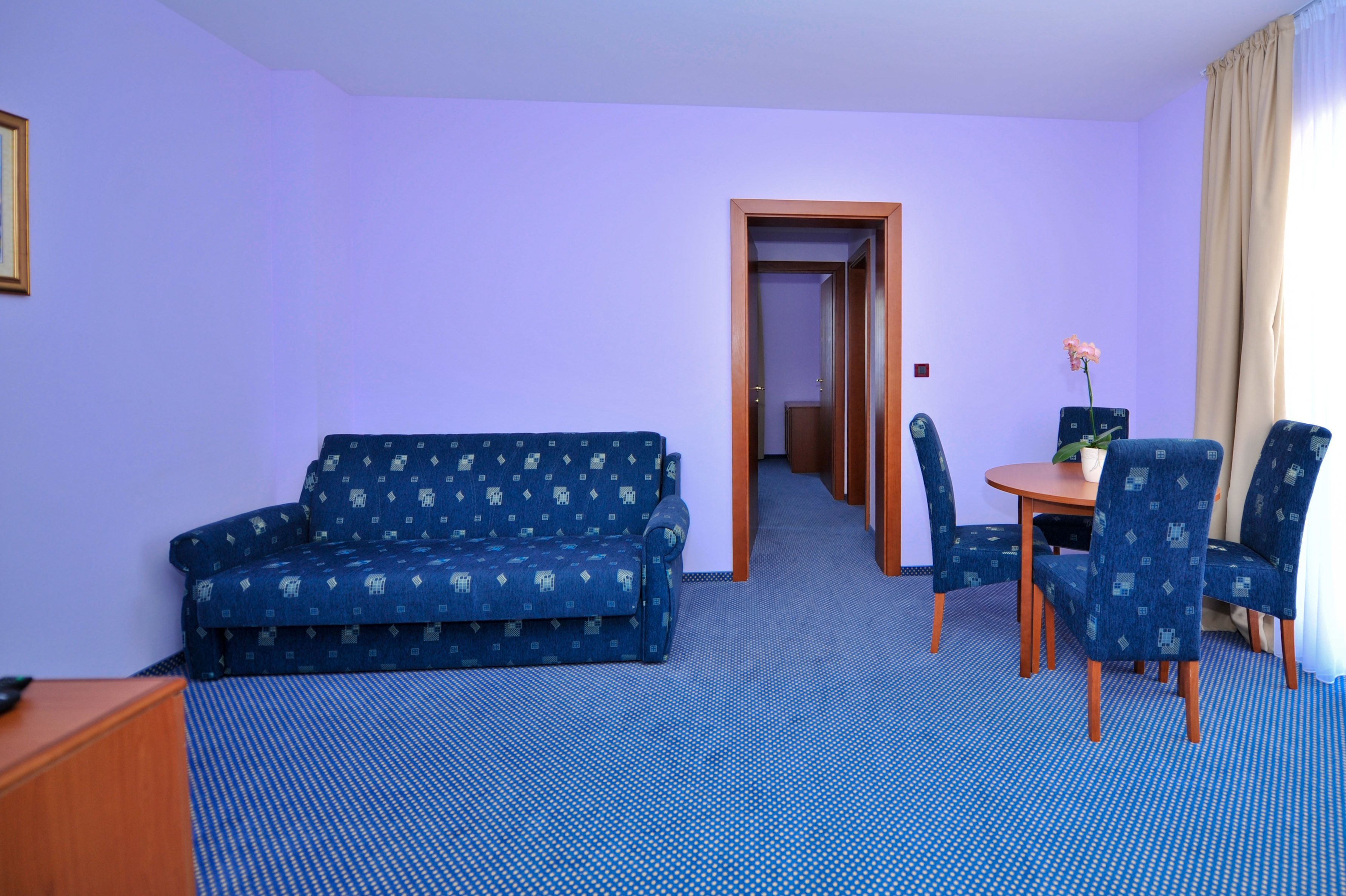 Quarto de hotel com um sofá azul estampado, uma mesa redonda pequena com duas cadeiras e uma porta de madeira que dá para outro espaço.