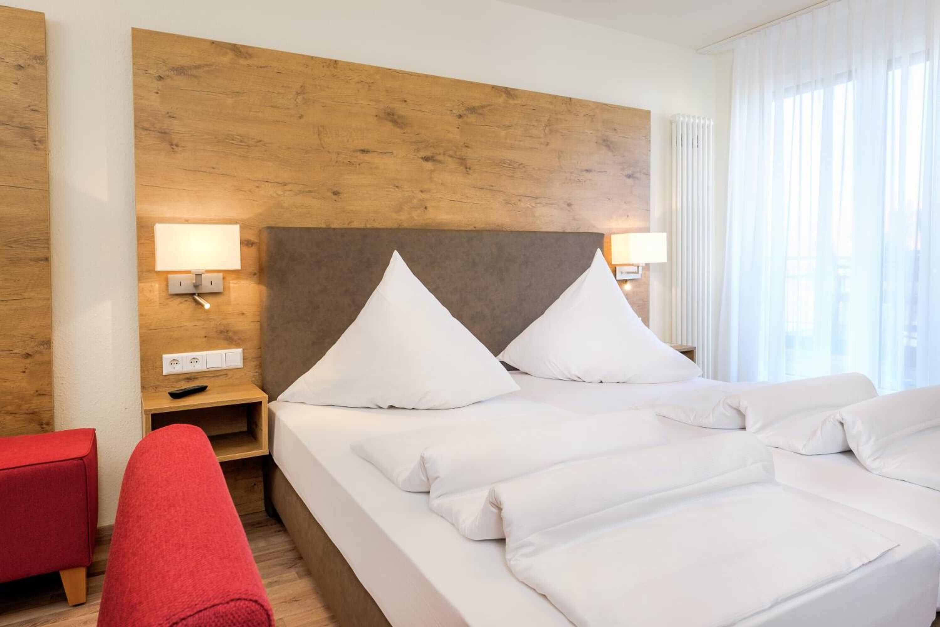 Quarto de hotel com cabeceira de madeira, duas luminárias de cabeceira, uma cadeira vermelha e uma cama grande com roupa de cama branca.