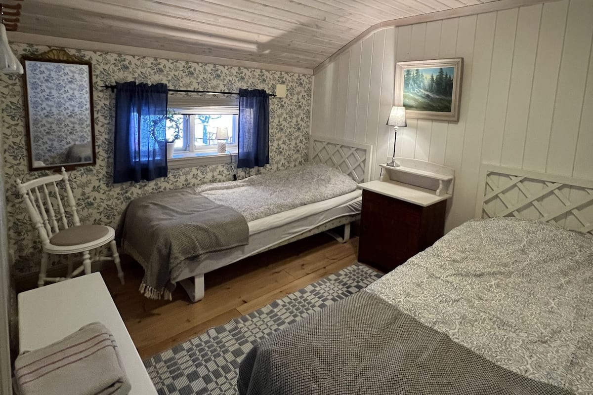 Quarto aconchegante com cama, escrivaninha e cadeira. Há uma janela com cortinas e um quadro na parede.