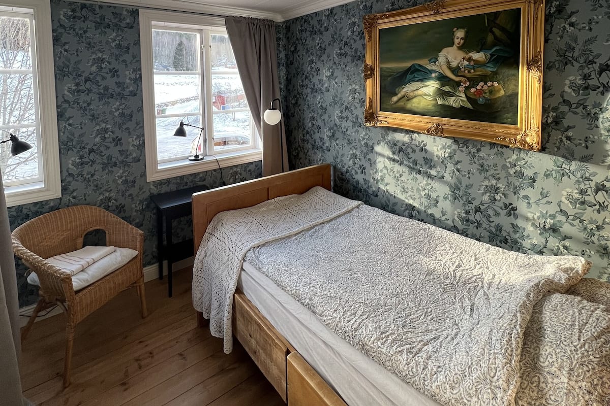 Quarto com cama de madeira, escrivaninha, cadeira de vime e um quadro na parede.