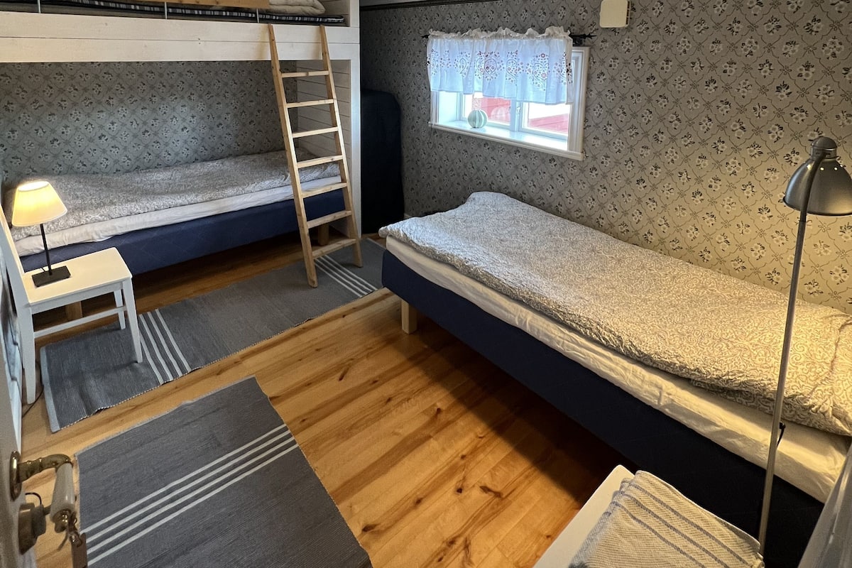 Quarto pequeno com beliches, uma escada, uma secretária compacta com abajur, janela com cortinas e piso de madeira.