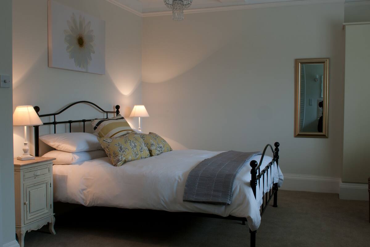 Uma cama bem arrumada com roupa de cama branca e almofadas florais, uma mesa de cabeceira com abajures e um espelho fixado na parede.
