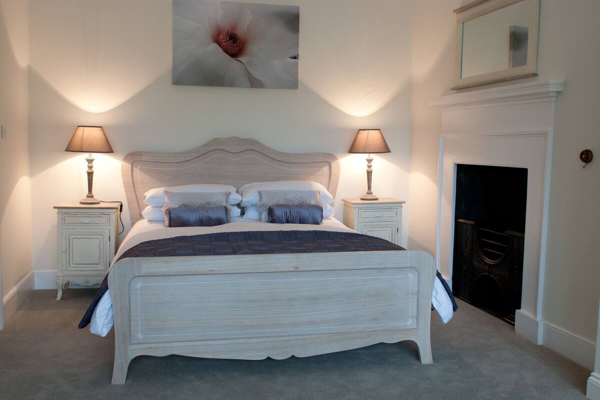 Quarto com uma cama grande, duas mesas de cabeceira com luminárias, uma lareira e um quadro floral na parede.