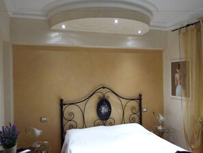 Quarto com cama, mesa de cabeceira, luminária, planta em vaso e um quadro na parede.