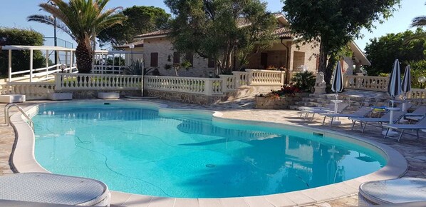Piscina com borda curva, rodeada por espreguiçadeiras e guarda-sóis, com um edifício de pedra ao fundo.