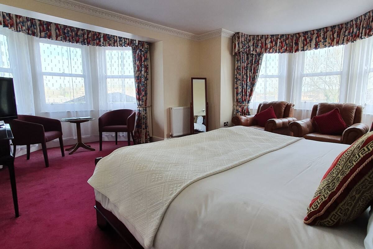 Quarto de hotel com uma cama grande, duas poltronas, uma escrivaninha com televisão e janelas com cortinas florais.